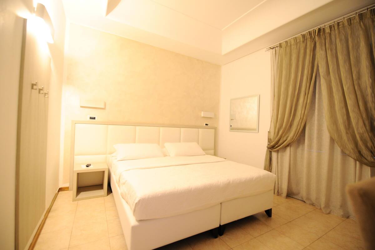 Quarto de hotel com uma cama grande, uma mesinha de cabeceira, uma luminária na parede e uma janela com cortinas.