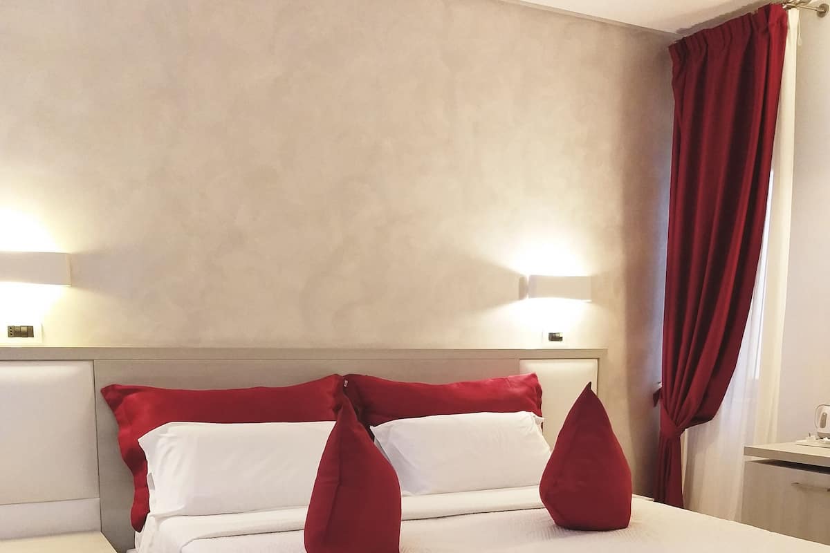 Cama bem arrumada com roupa de cama branca e vermelha, mesa de cabeceira e luminária na parede.
