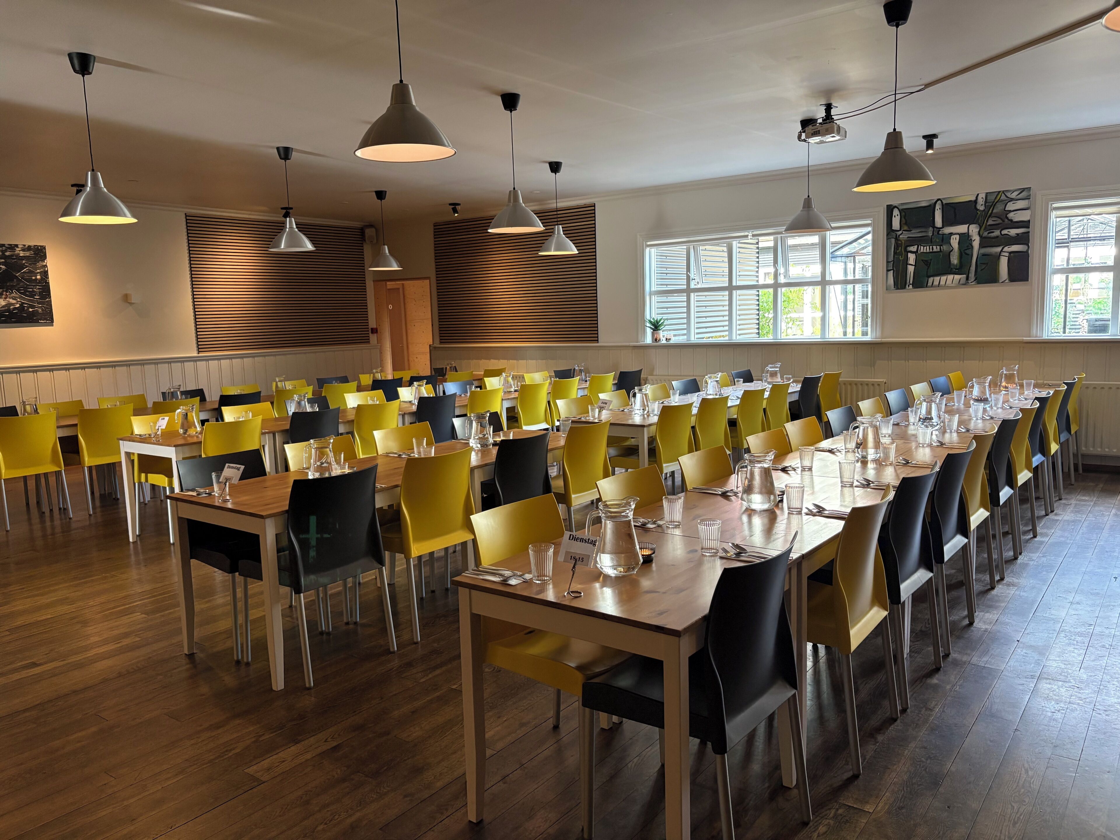 Uma sala de jantar ampla, com várias mesas e cadeiras, janelas grandes e iluminação pendente.