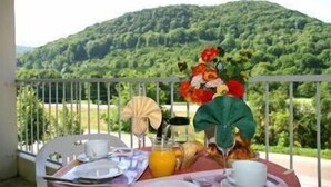 Uma varanda com uma mesa posta para o café da manhã, um vaso de flores e vista para uma colina arborizada.