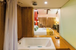 Quarto de hotel com piso de madeira, uma cama grande com colcha amarela, uma escrivaninha com abajur e um banheiro com banheira.