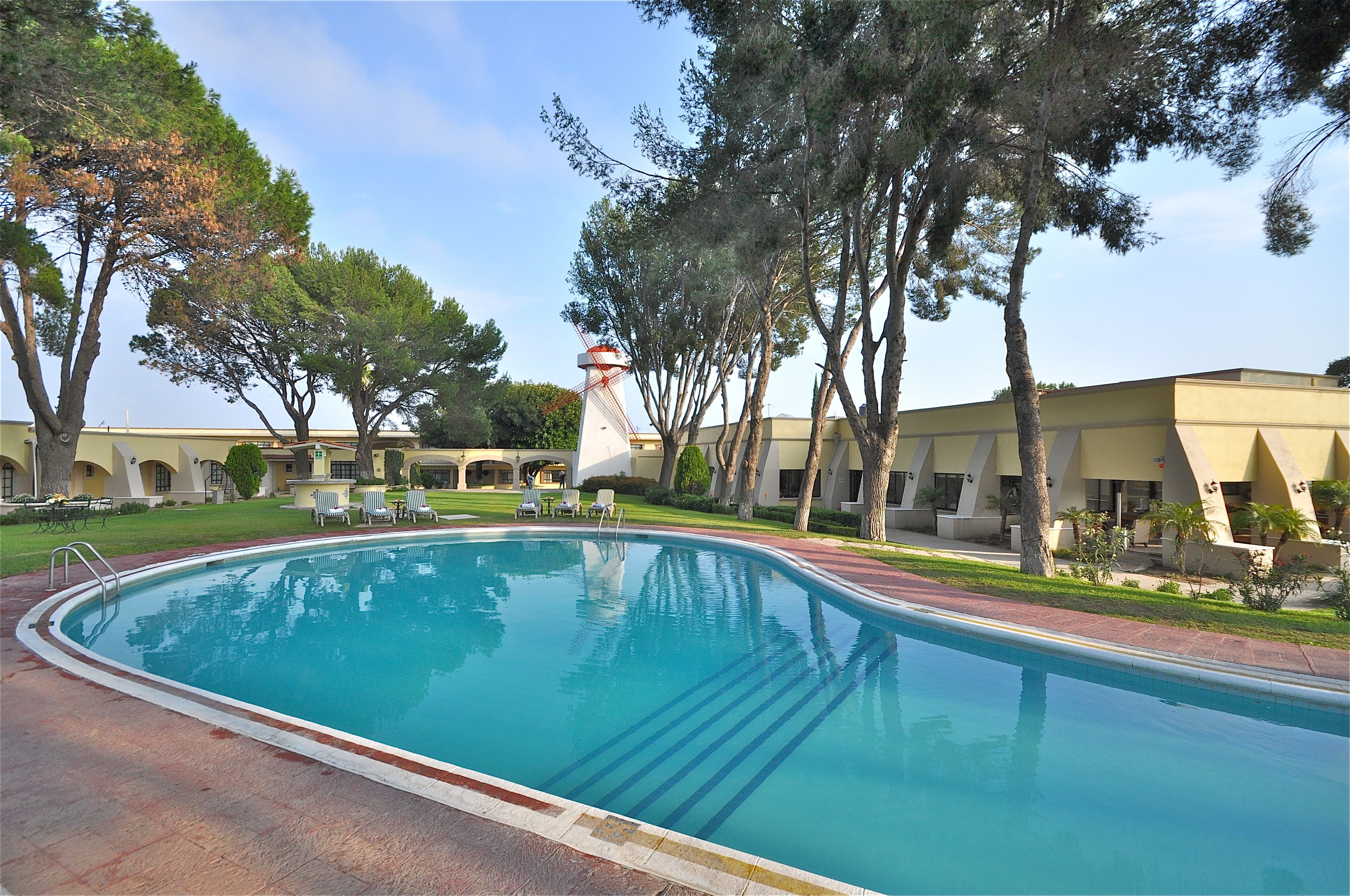 Piscina com deck revestido, cercada por árvores e um edifício com fachada clara.