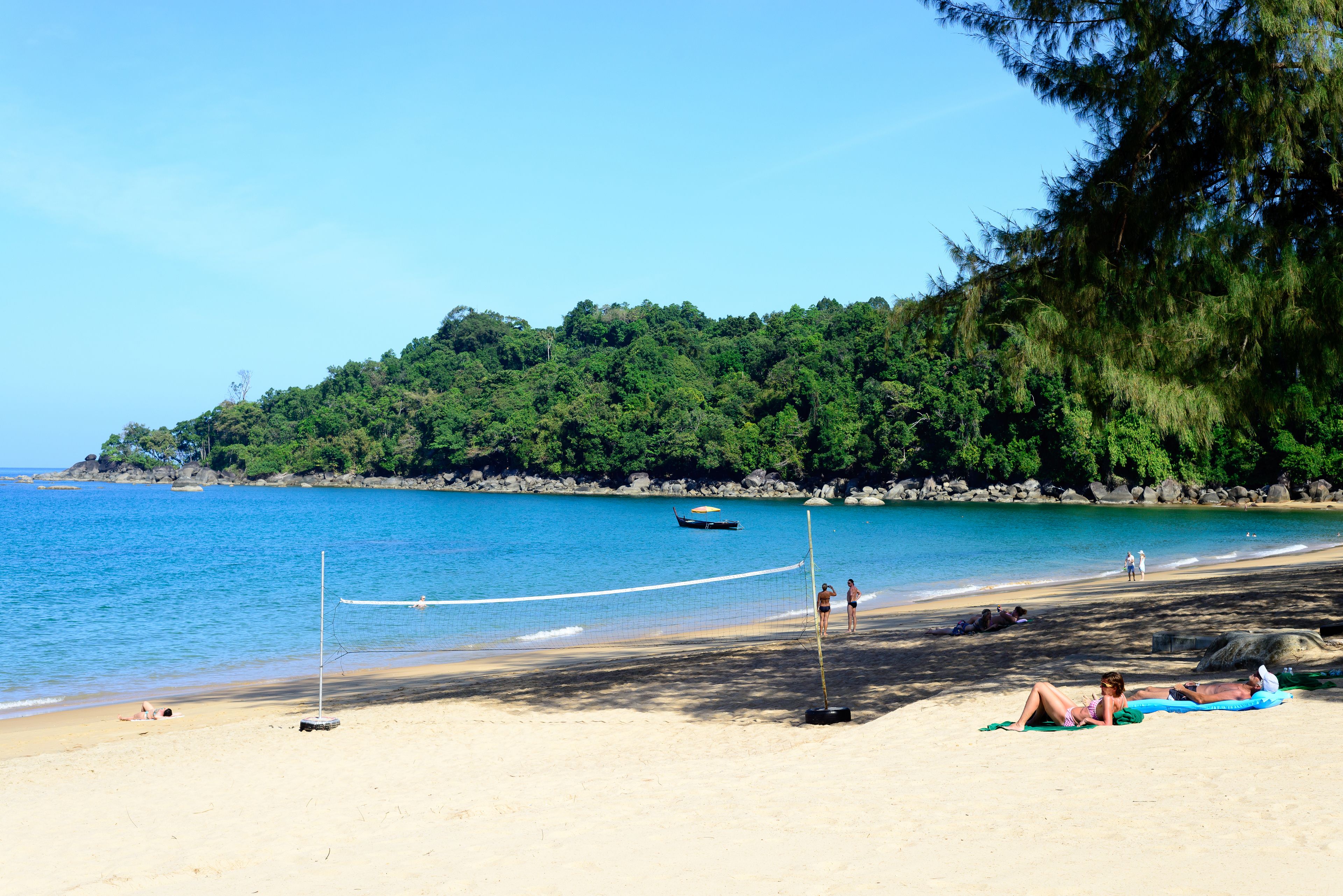 Uma praia com uma rede de voleibol, pessoas tomando sol e um barco na água.
