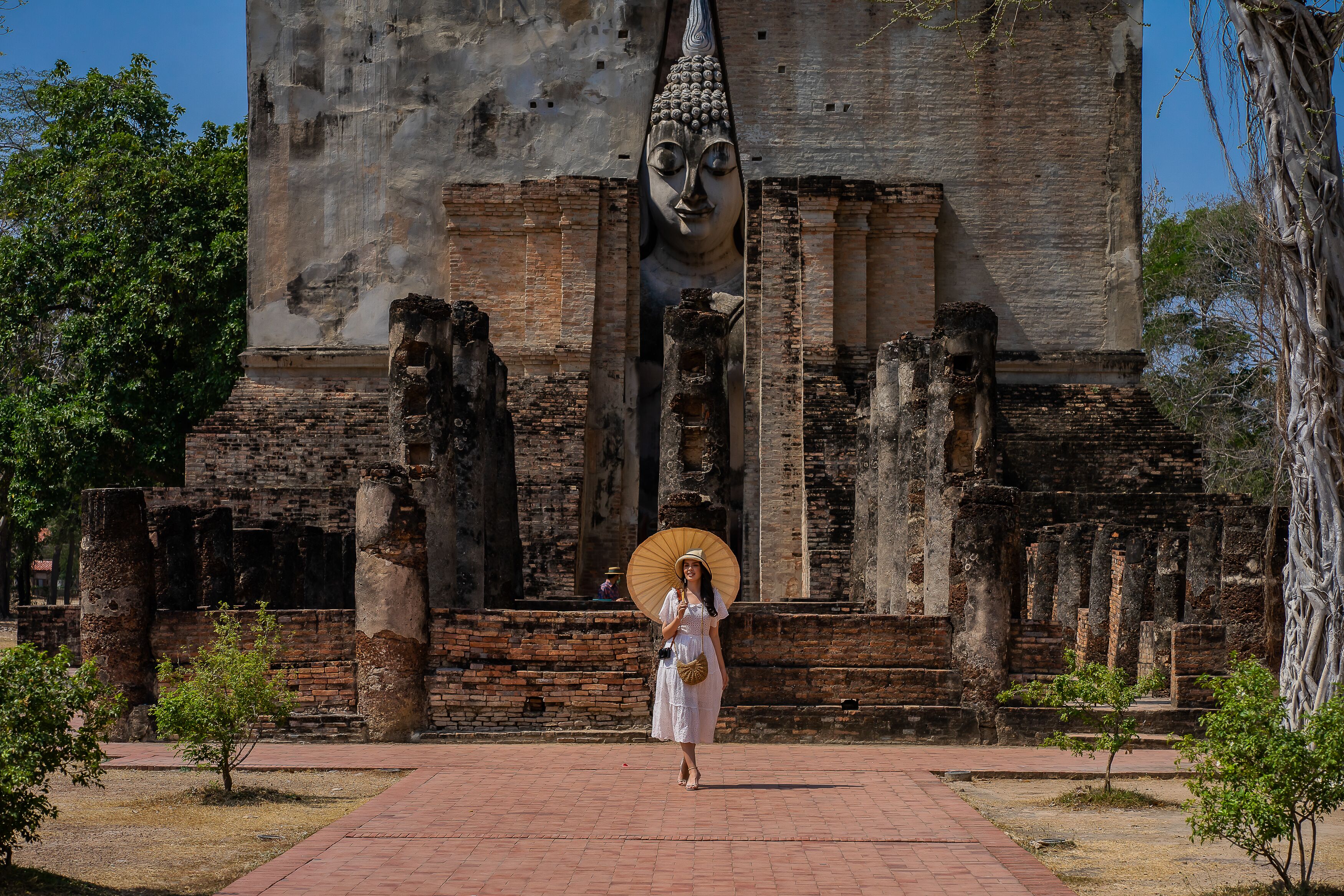 Uma mulher de vestido branco em frente a uma grande estátua de Buda, com ruínas antigas de tijolo ao fundo.
