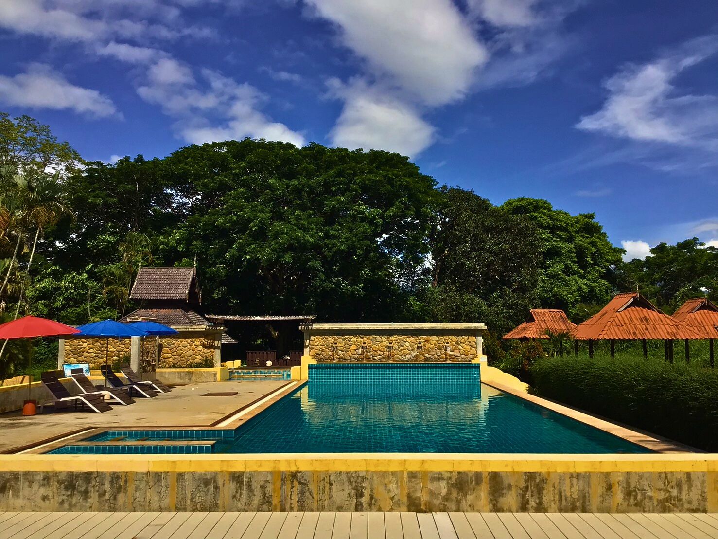 Uma piscina com espreguiçadeiras, um muro de pedra e uma construção com telhado de palha.