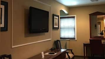 Quarto com televisão na parede, escrivaninha com cadeira, cómoda e janela com persianas.