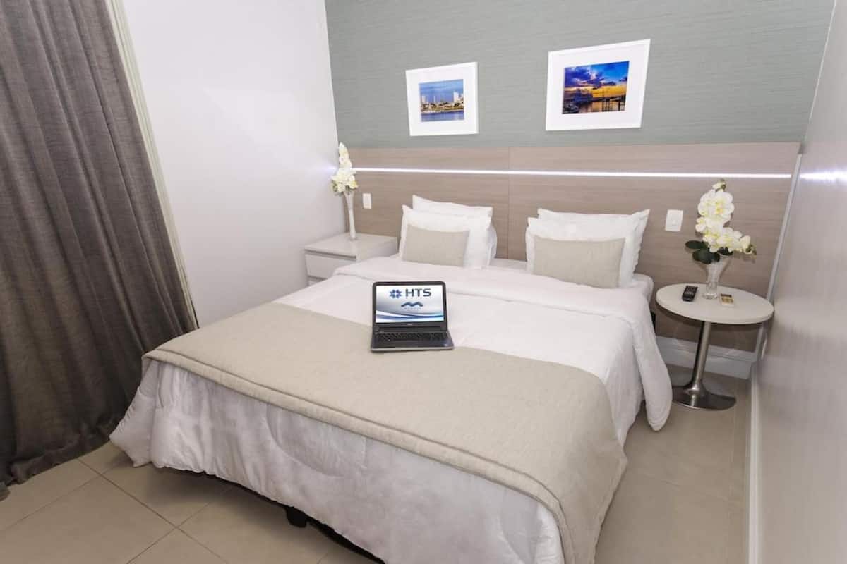 Quarto de hotel com uma cama, mesa de cabeceira com abajur, uma mesinha redonda e duas imagens emolduradas na parede.