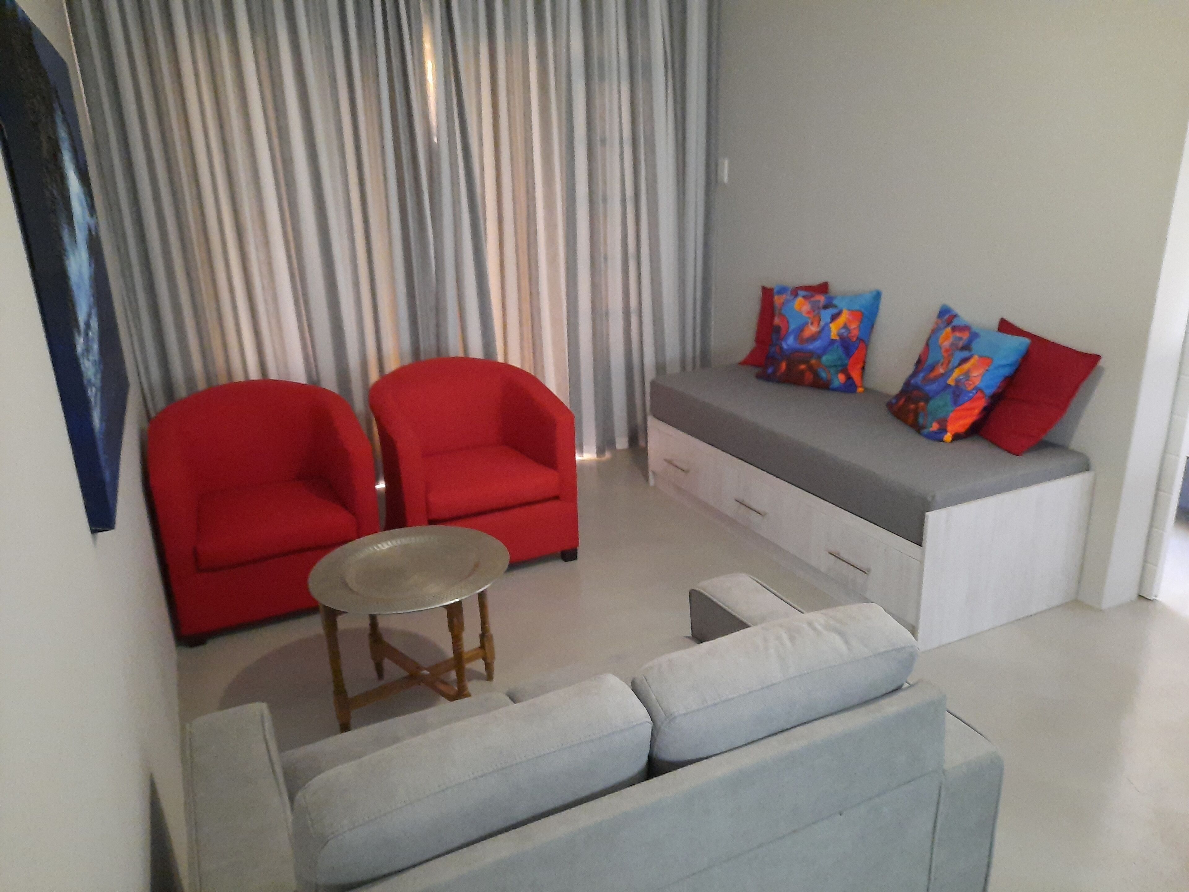 Uma sala de estar moderna com um sofá cinza, duas poltronas vermelhas, uma mesa redonda pequena e um armário branco com almofadas coloridas.