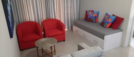Uma sala de estar moderna com um sofá cinza, duas poltronas vermelhas, uma mesa redonda pequena e um armário branco com almofadas coloridas.
