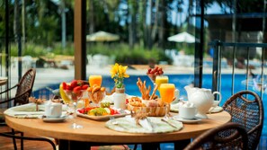 Mesa de jantar posta com itens de café da manhã, vista para a piscina e mobiliário de exterior.