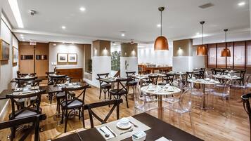 Uma sala de jantar moderna com várias mesas e cadeiras, algumas com cadeiras de plástico transparente e outras com cadeiras de madeira escura. Há luminárias pendentes no teto, e uma placa indicando uma área 'SAIDA'.