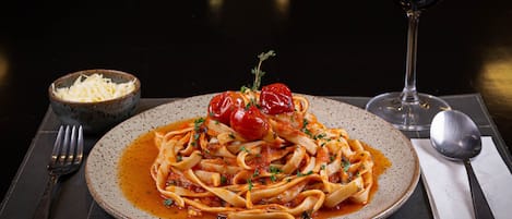 Um prato de massa com tomate cereja e ervas, um copo de vinho tinto e uma tigela de queijo ralado em cima de uma mesa.