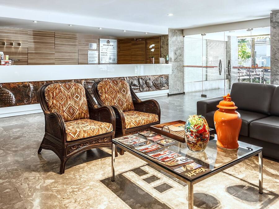 Um lobby com balcão de recepção, duas poltronas estampadas, um sofá, uma mesa de centro de vidro com revistas e um vaso com flores.