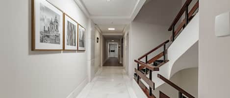 Um corredor com quadros nas paredes, escada com corrimão de madeira e piso ladrilhado.