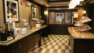 Uma cozinha de hotel com chão xadrez, equipada com máquinas de café, um sistema de buffet e diversos eletrodomésticos.