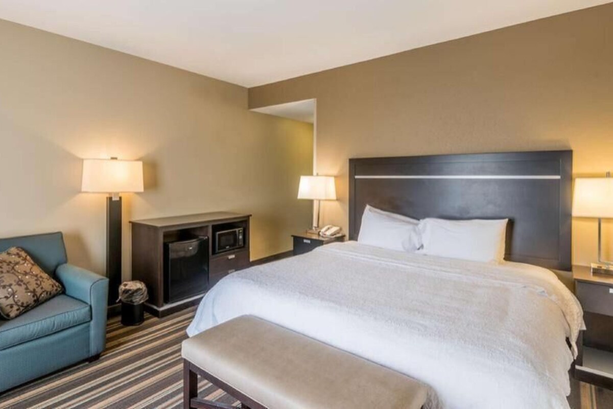 Quarto de hotel com cama, poltrona, escrivaninha, luminária e telefone.