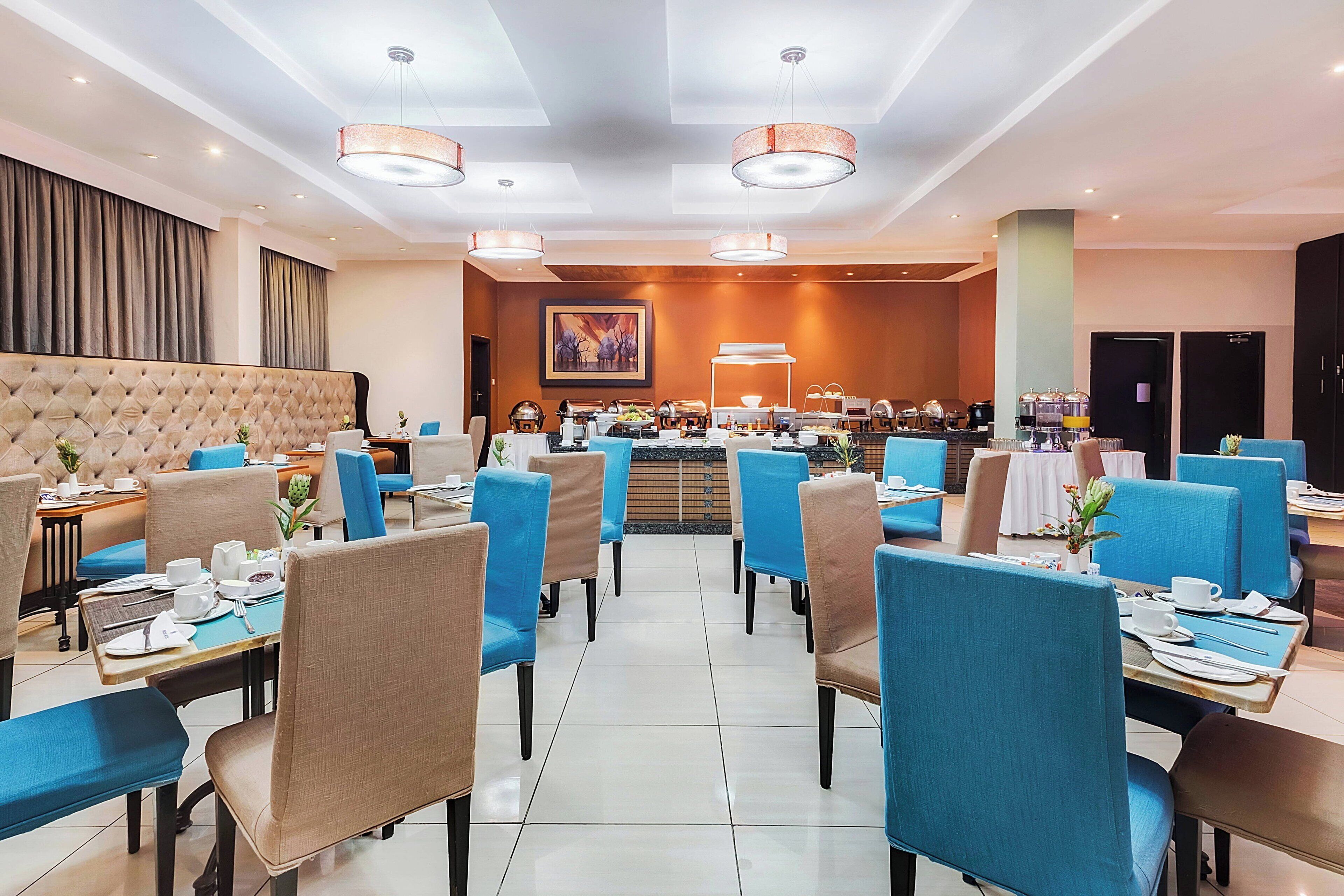 Restaurante com cadeiras azuis e bege, uma grande cabeceira estofada e um balcão central com diversos itens.