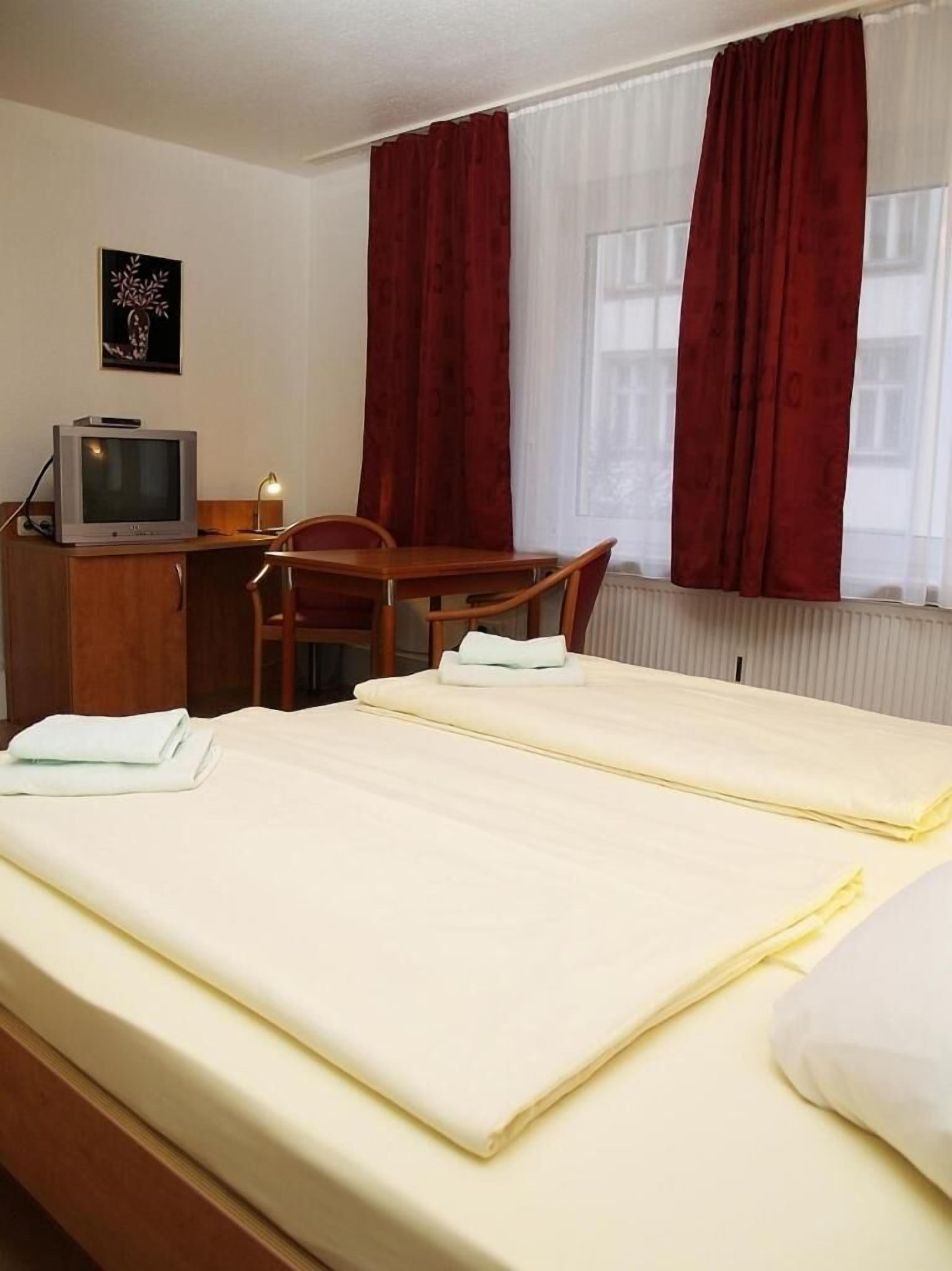 Quarto de hotel com duas camas, uma escrivaninha com televisão, uma cadeira e cortinas vermelhas.