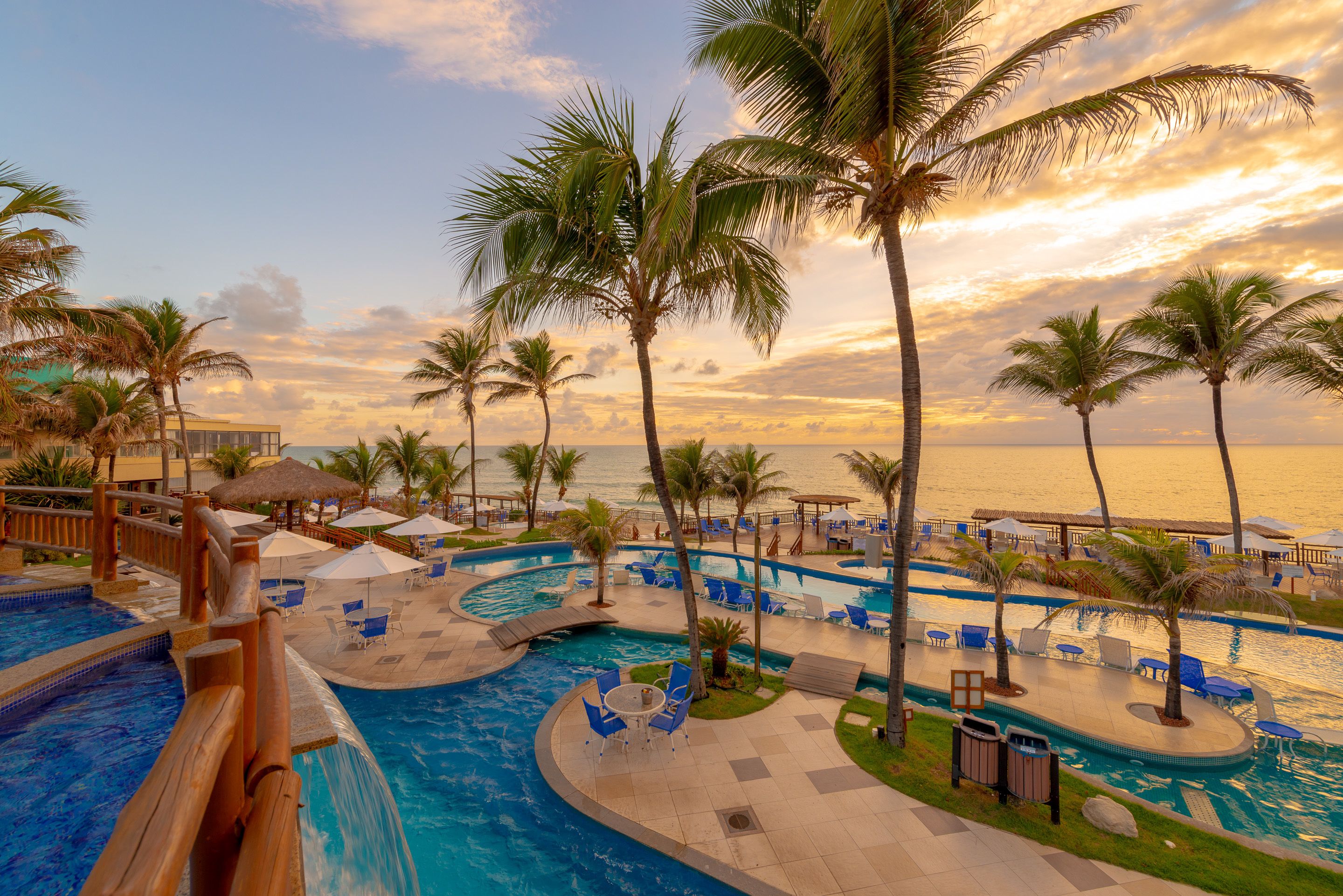 Área de piscina do resort com palmeiras, espreguiçadeiras e vista para o oceano ao pôr do sol.