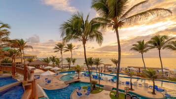 Área de piscina do resort com palmeiras, espreguiçadeiras e vista para o oceano ao pôr do sol.