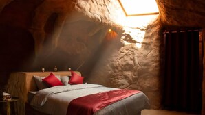 Quarto com parede de pedra, cama com roupa de cama vermelha e cinza, e um claraboia.