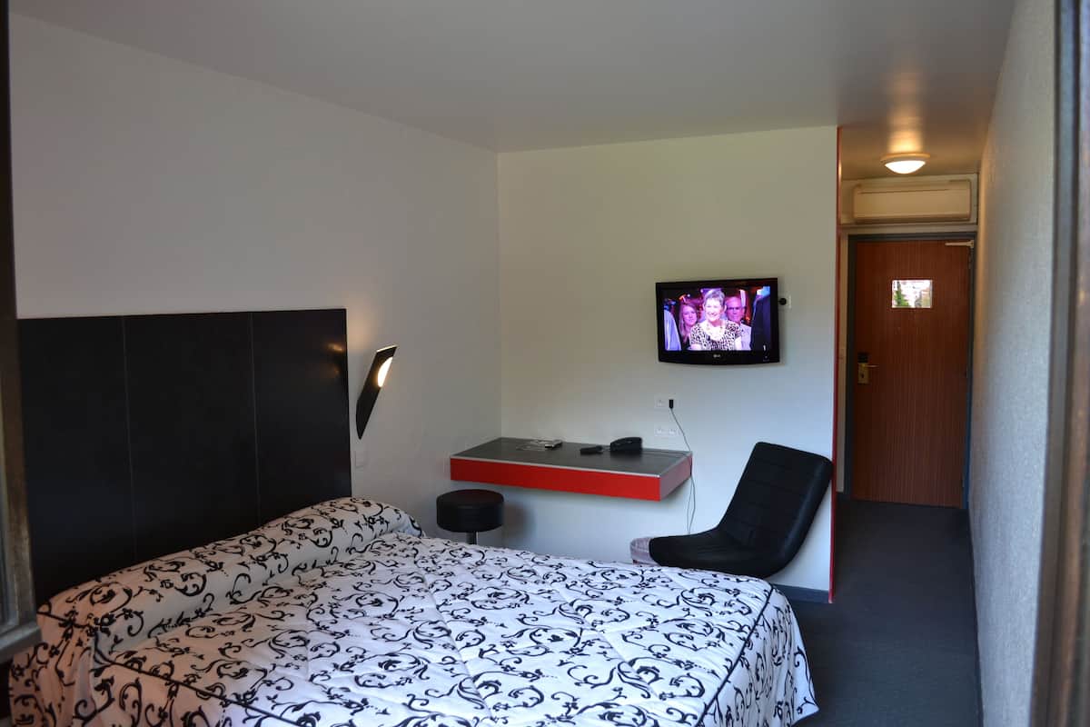 Quarto de hotel com cama, televisão fixada na parede, pequena escrivaninha e uma cadeira.