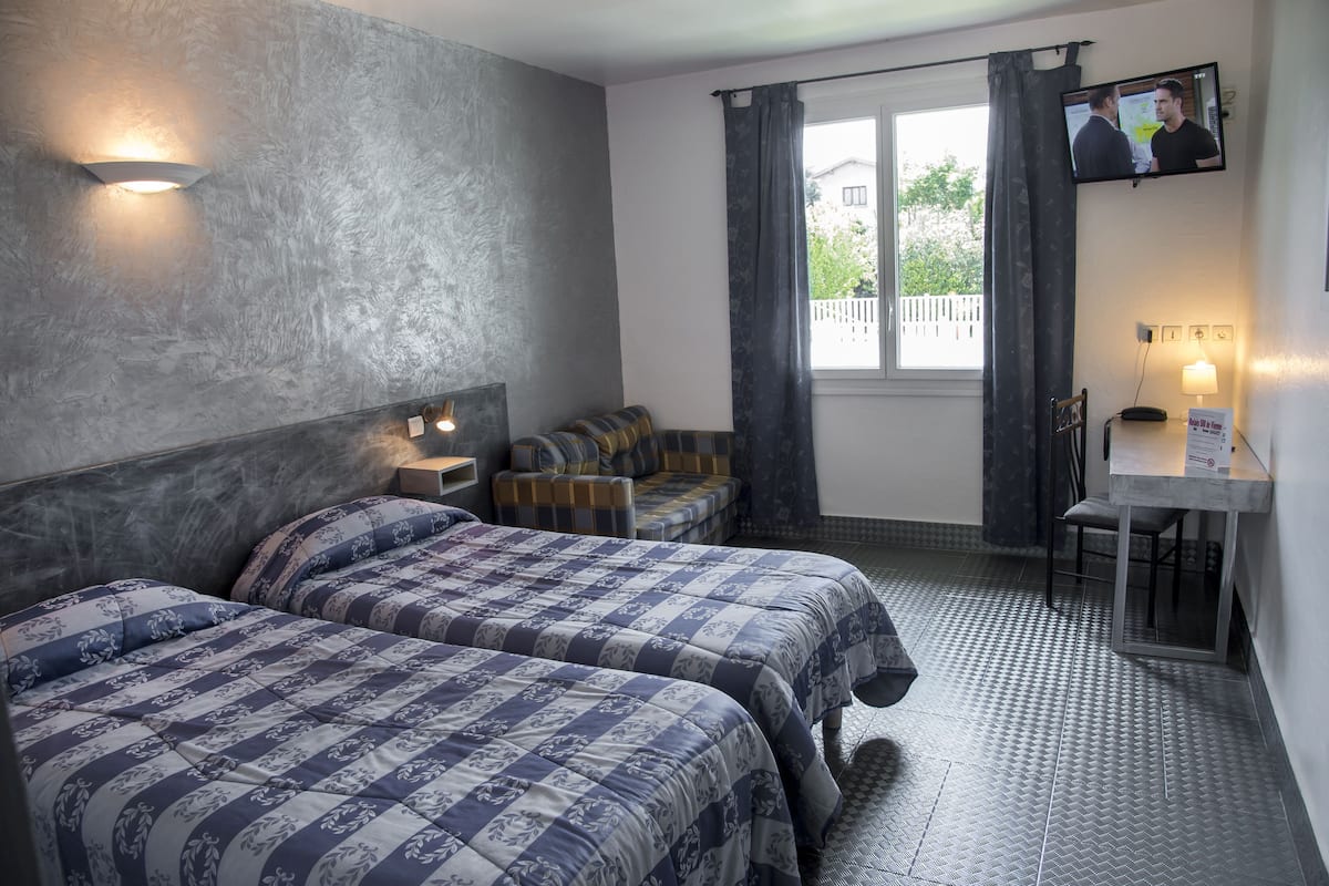 Quarto de hotel com duas camas, um sofá pequeno, uma escrivaninha e televisão na parede.
