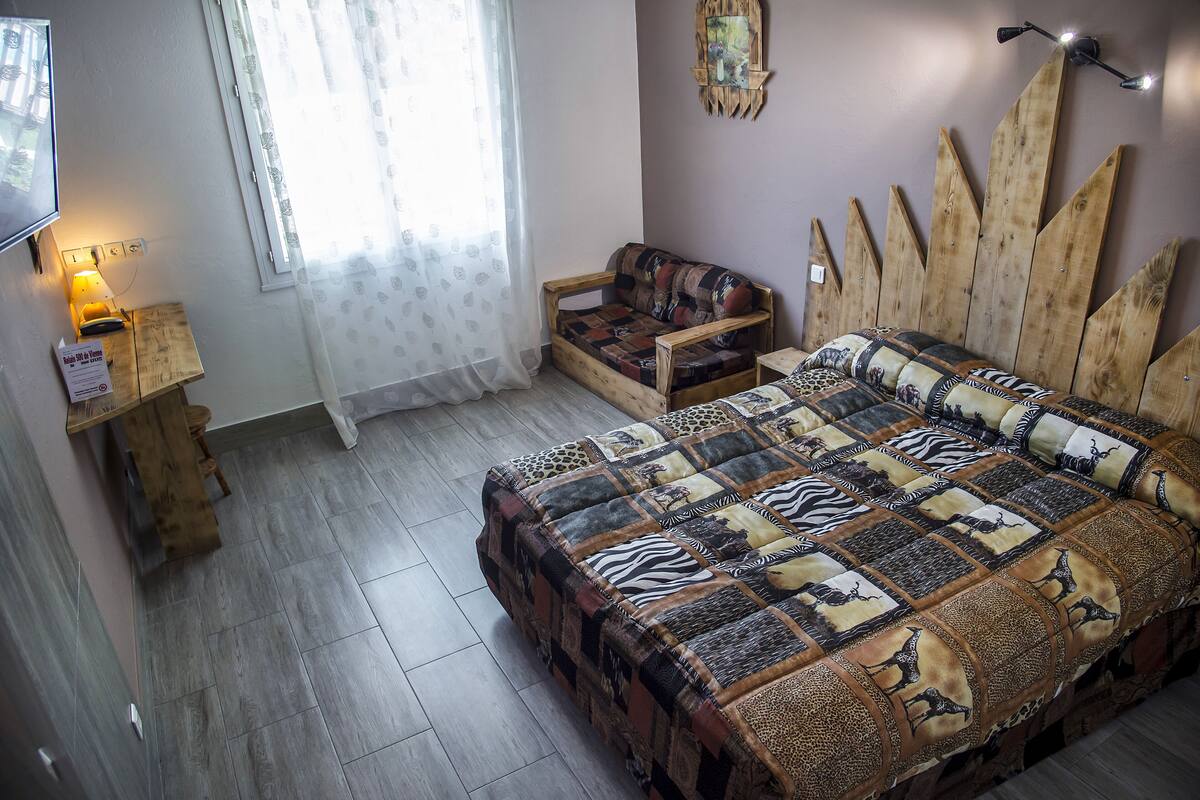 Quarto com cabeceira de madeira, colcha acolchoada, um pequeno banco de madeira e luminária fixada na parede.