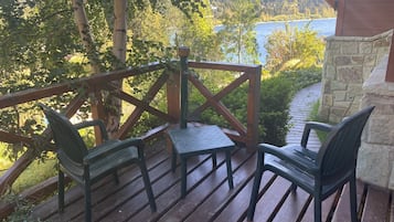 Um deck de madeira com duas cadeiras e uma mesa, com vista para um lago e área arborizada.