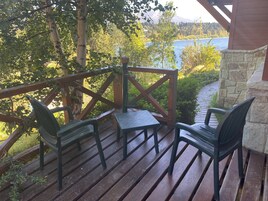 Um deck de madeira com duas cadeiras e uma mesa, com vista para um lago e área arborizada.