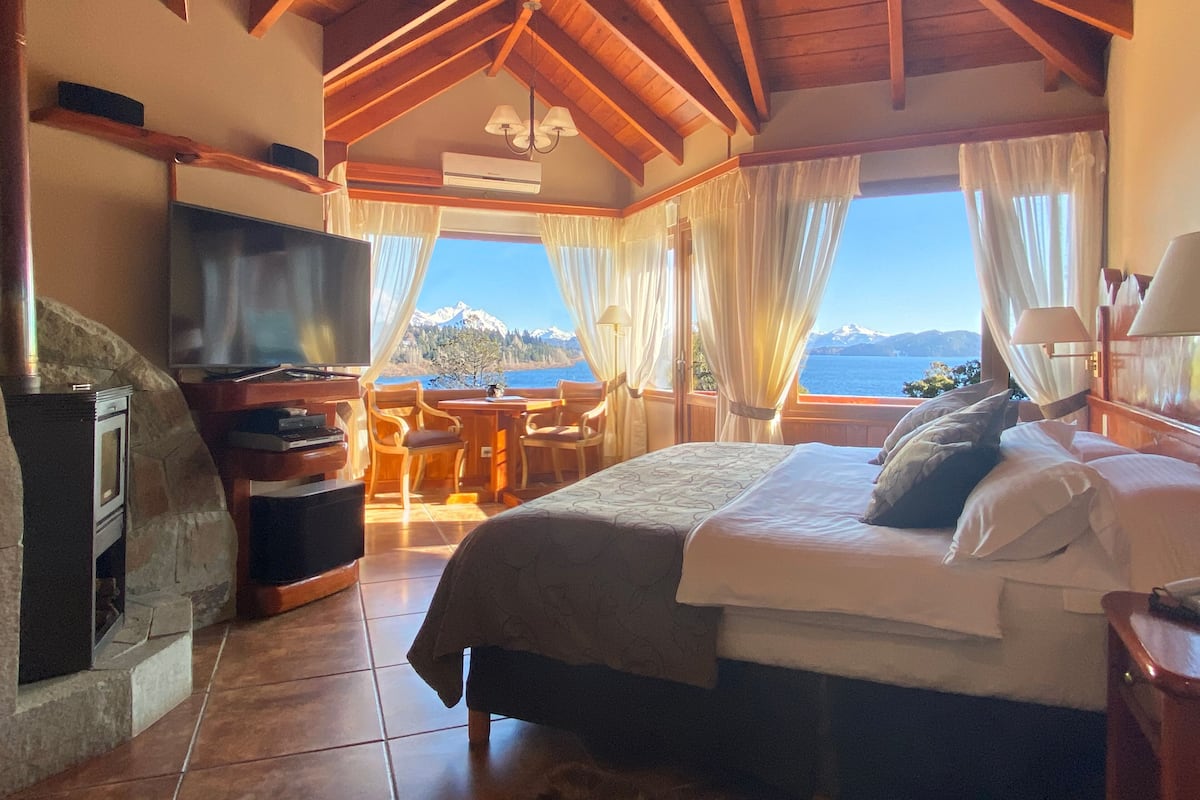 Quarto com cama, televisão, lareira e vista para montanhas e um lago.