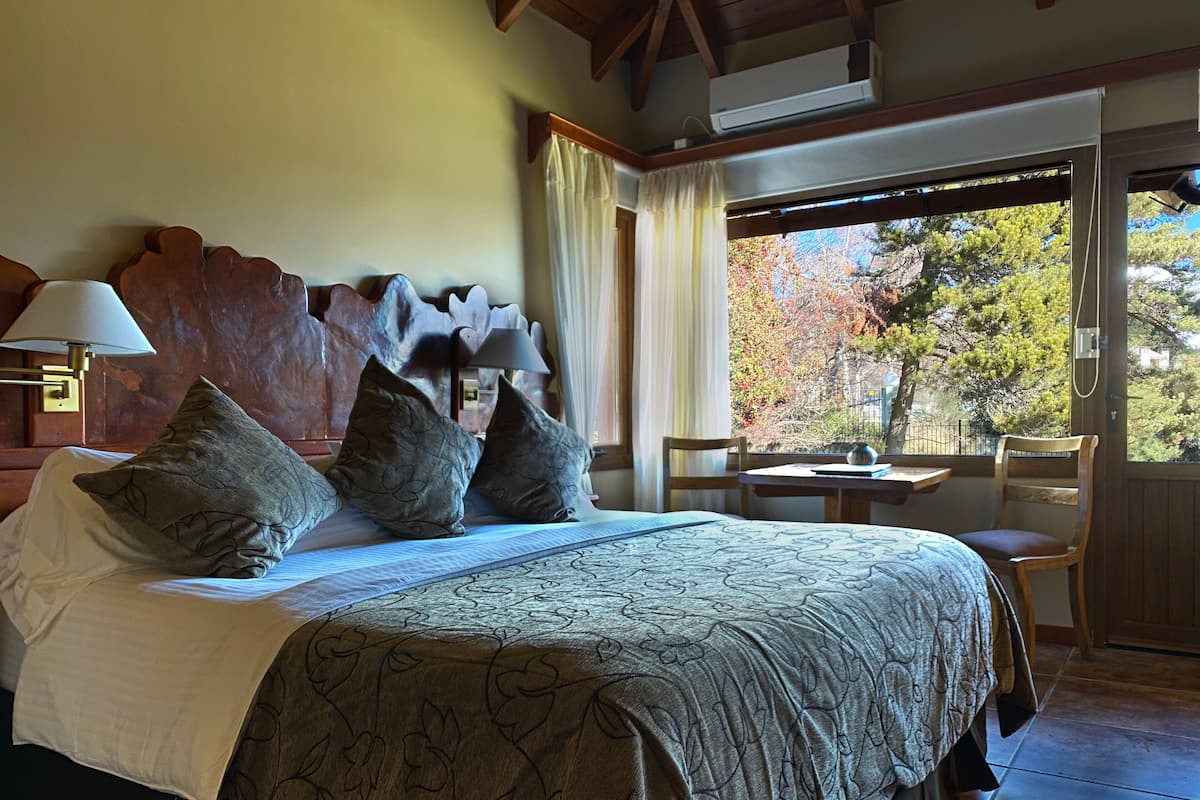 Quarto de hotel com uma cama grande, cabeceira de madeira, uma mesa com uma cadeira e vista para árvores através de uma porta de vidro.