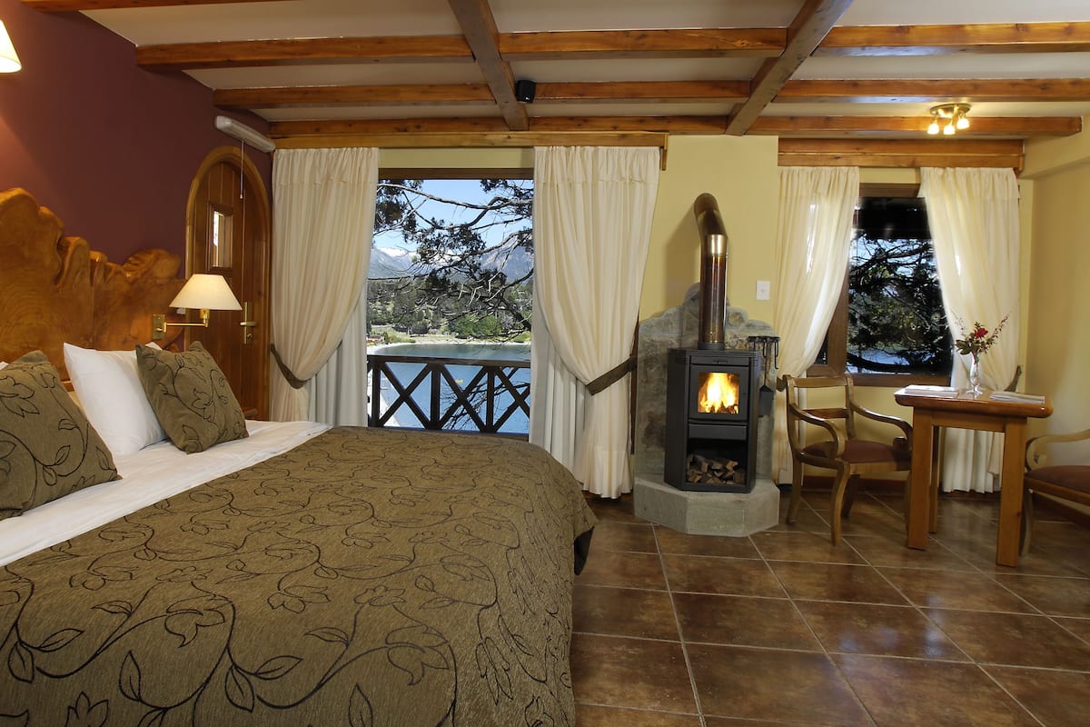 Quarto com cama, cabeceira de madeira, mesa de cabeceira com abajur, lareira de pedra, vista para o exterior e uma pequena mesa com um vaso.