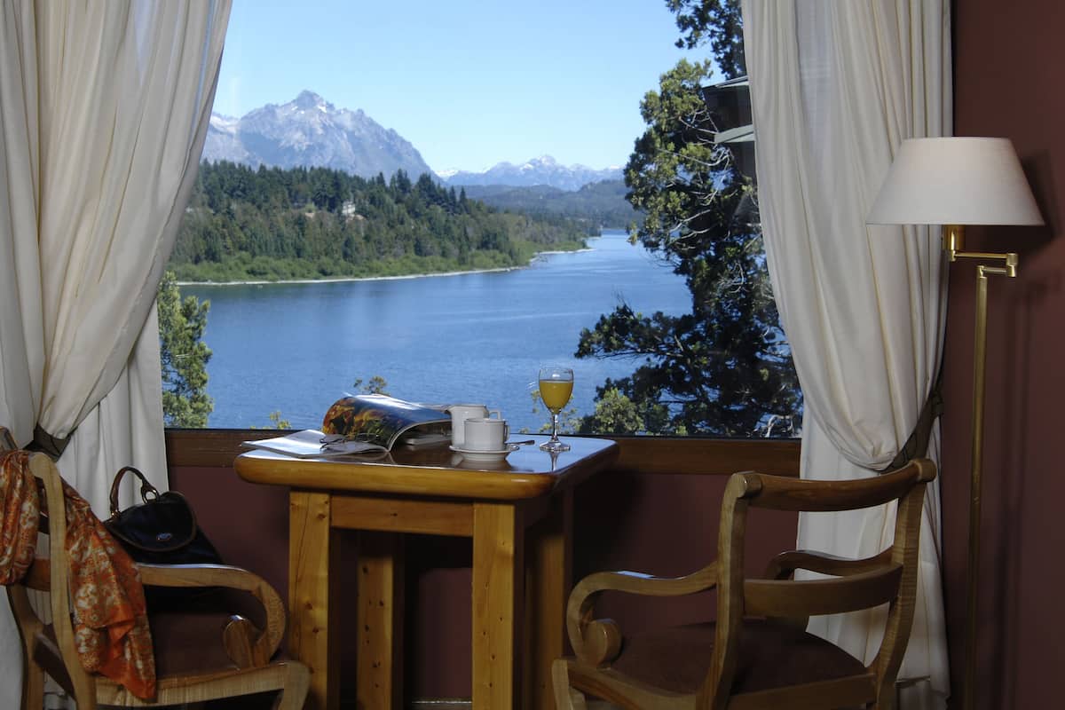 Quarto com uma mesa posta para dois, uma luminária e vista para um lago com montanhas ao fundo.