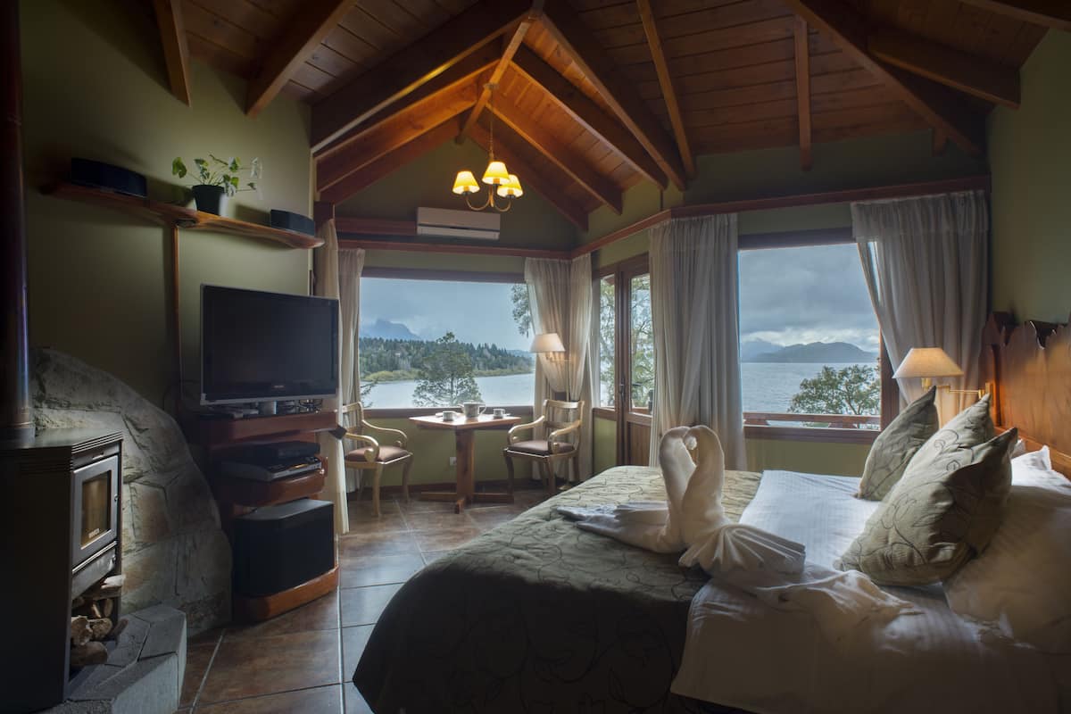 Um quarto aconchegante com lareira, televisão, cama com almofadas em formato de cisne, mesa de jantar e vista para um lago e montanhas.