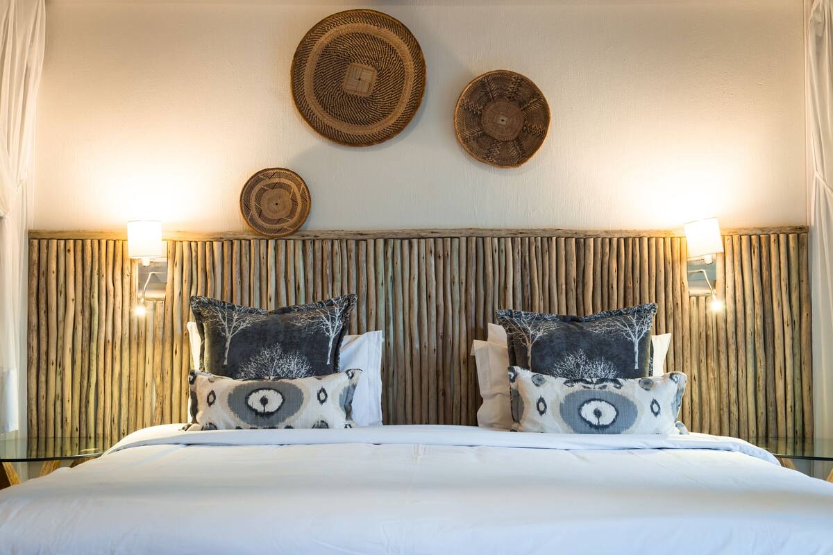 Cama com almofadas estampadas, cabeceira de madeira e luminárias fixadas na parede.