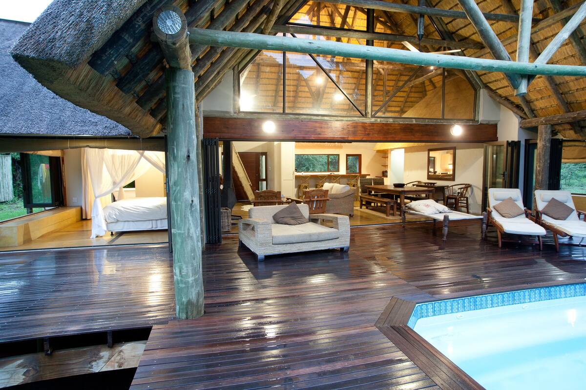 Um espaço interno amplo com deck de madeira, piscina e área de refeições com várias cadeiras e mesas.
