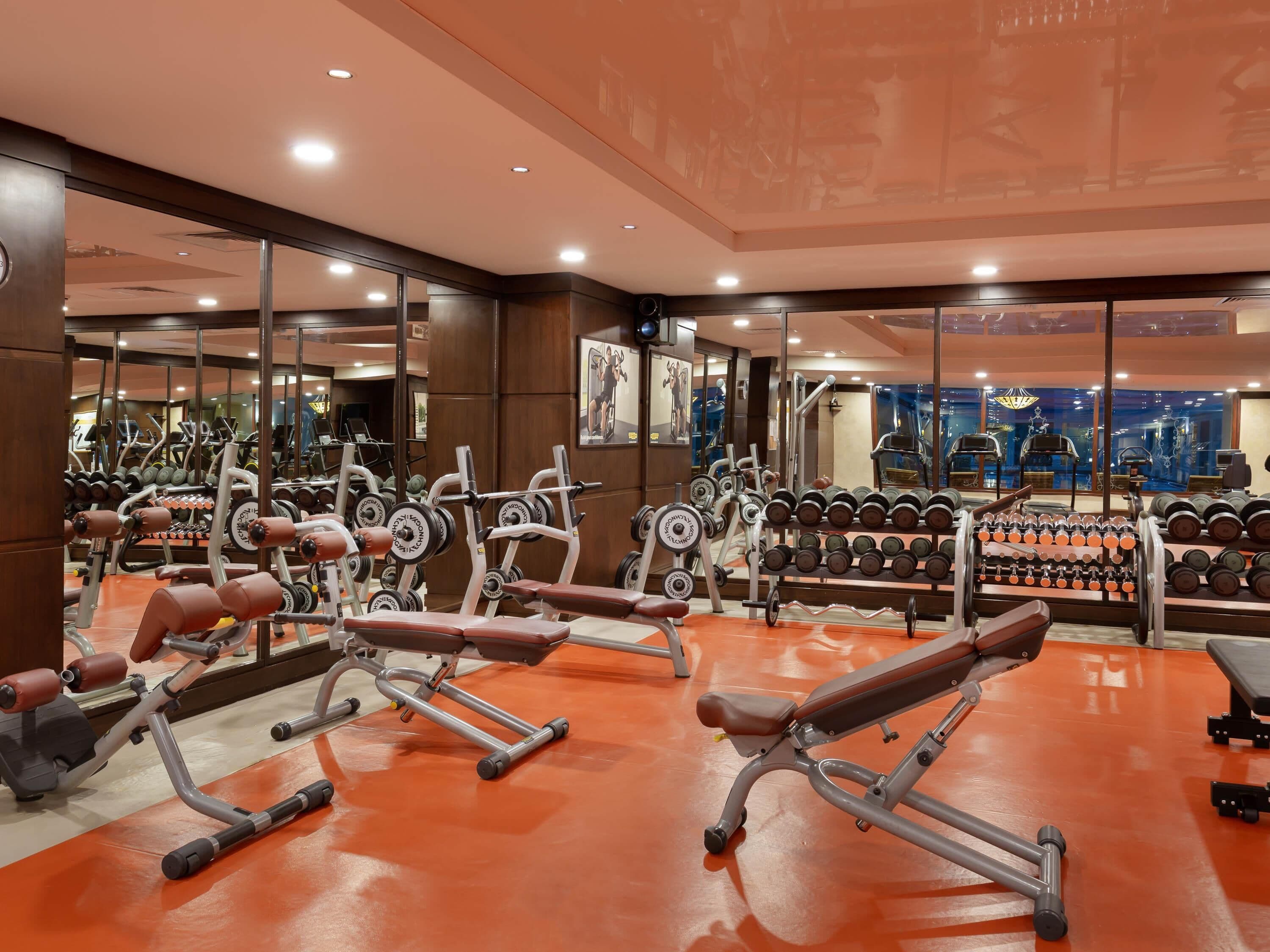 Academia com diversos equipamentos de exercício, incluindo bancos e halteres, e um espelho refletindo os aparelhos.
