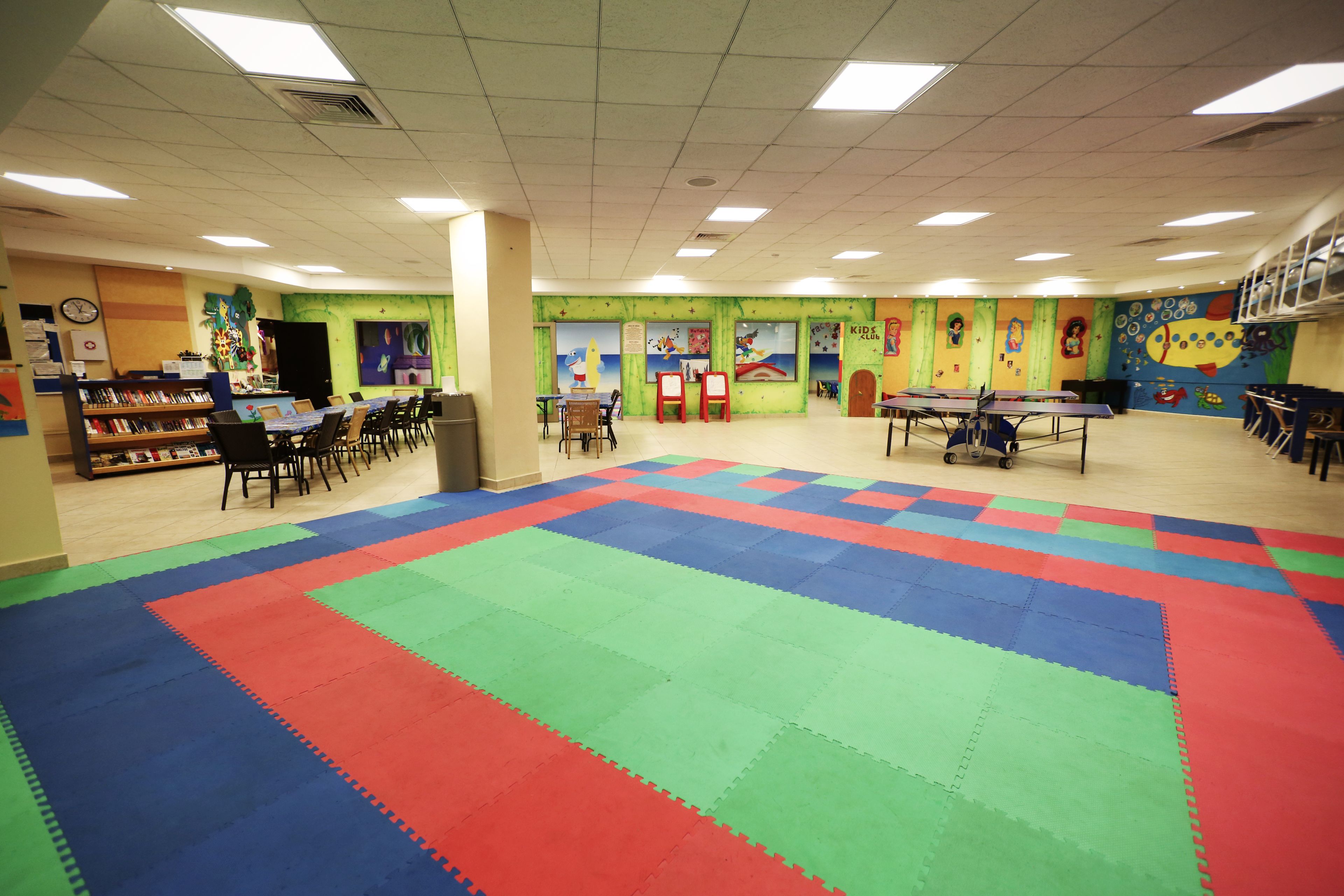Espaço interior bem iluminado com área de brincadeiras colorida, mesas, cadeiras e painéis educativos.