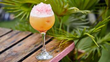 Um cocktail num copo sobre uma mesa de madeira, com uma planta verde ao fundo.