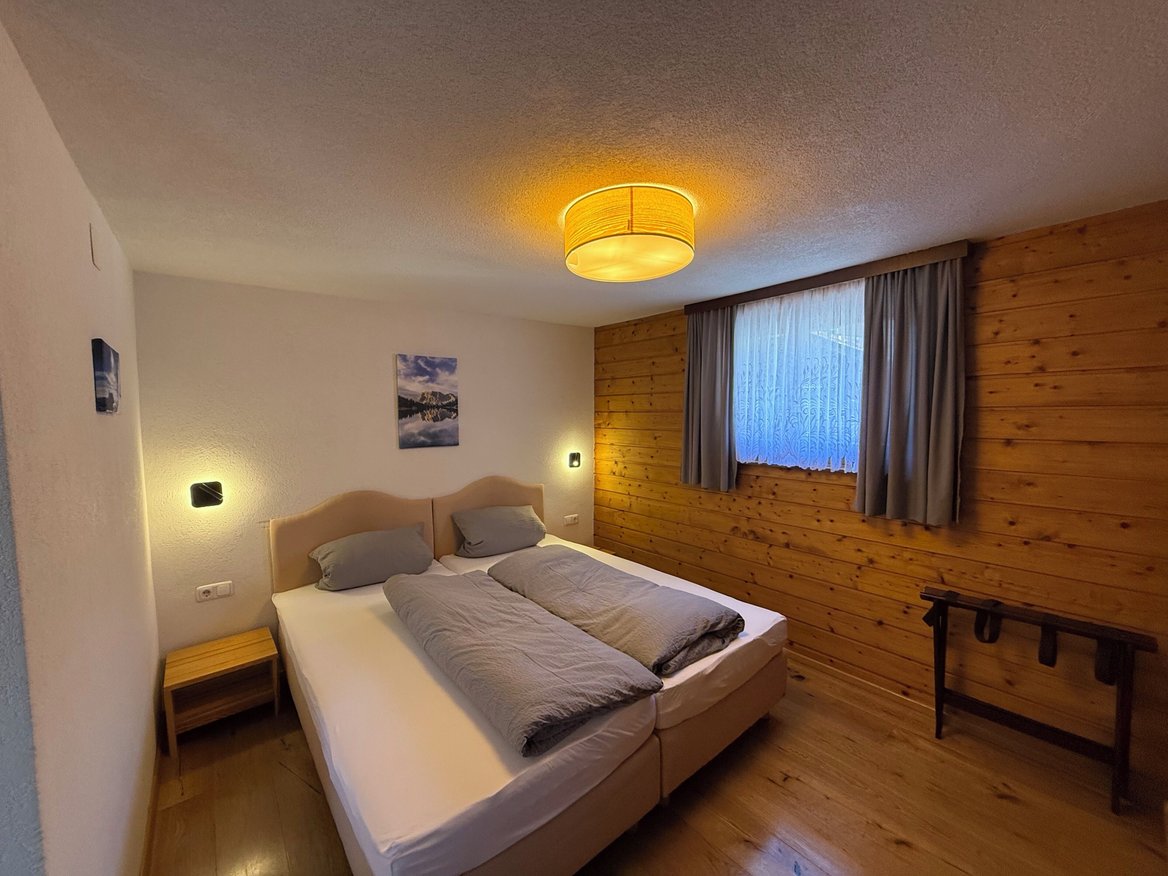 Quarto com parede de madeira, uma cama com almofadas, uma mesa de cabeceira, um lustre e uma janela com cortinas.
