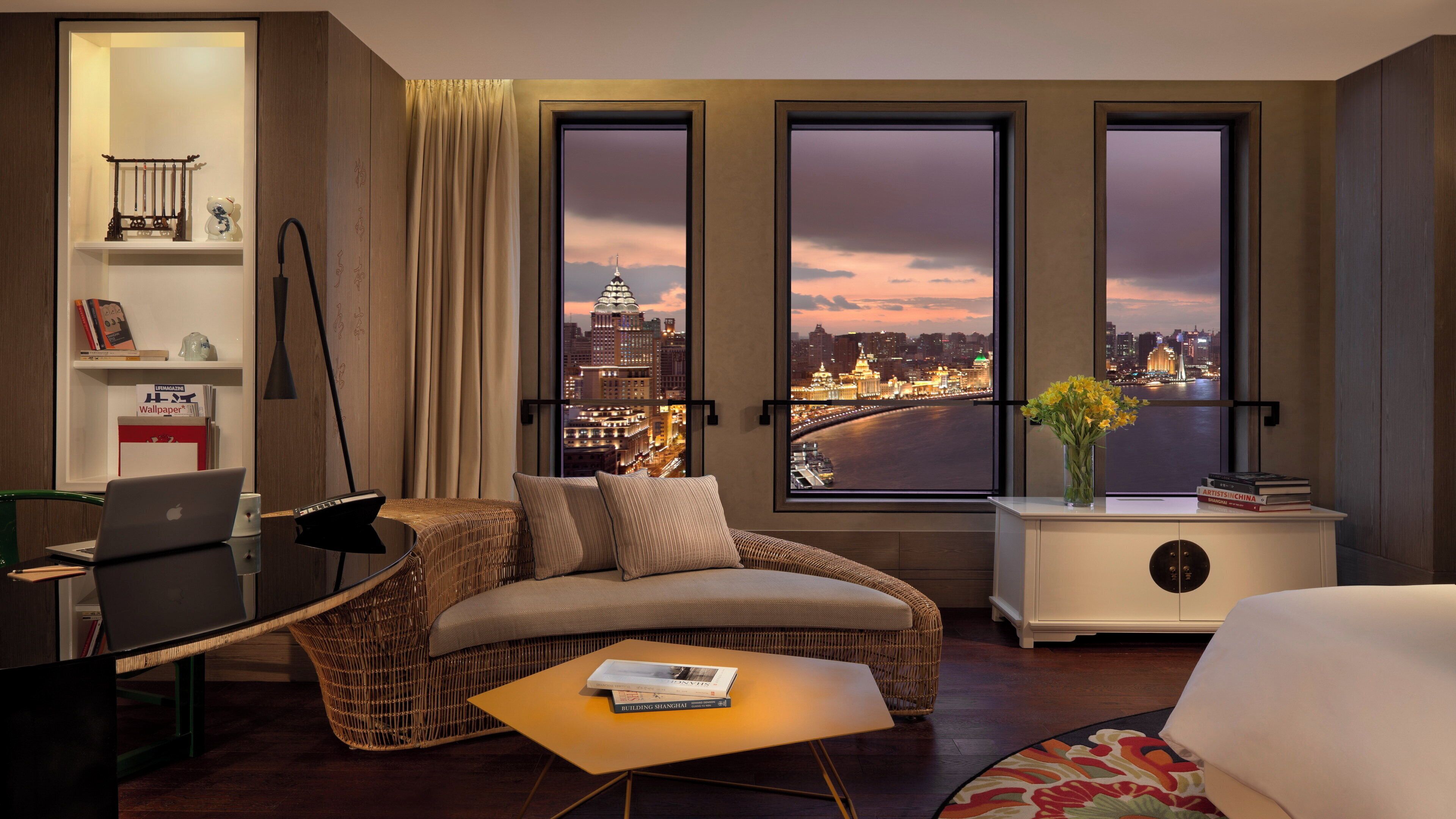 Quarto de hotel com vista para o horizonte da cidade, um sofá, uma escrivaninha com um laptop e uma mesinha com livros.