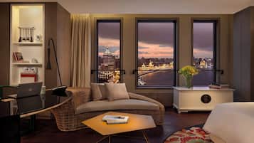 Quarto de hotel com vista para o horizonte da cidade, um sofá, uma escrivaninha com um laptop e uma mesinha com livros.