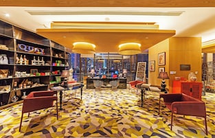 Um hall de hotel com design moderno, destacando uma grande estante de livros, um balcão de recepção e um tapete estampado.