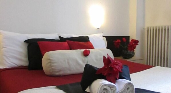 Quarto de hotel com cama bem arrumada, roupa de cama vermelha e branca e um vaso com flores vermelhas na mesa de cabeceira.
