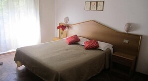 Cama bem arrumada com colcha marrom e duas almofadas vermelhas, mesa de cabeceira de madeira com um vaso de flores vermelhas e um relógio de parede.