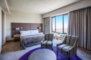 Quarto de hotel com uma cama grande, duas poltronas, uma mesa de cabeceira e vista para a cidade.