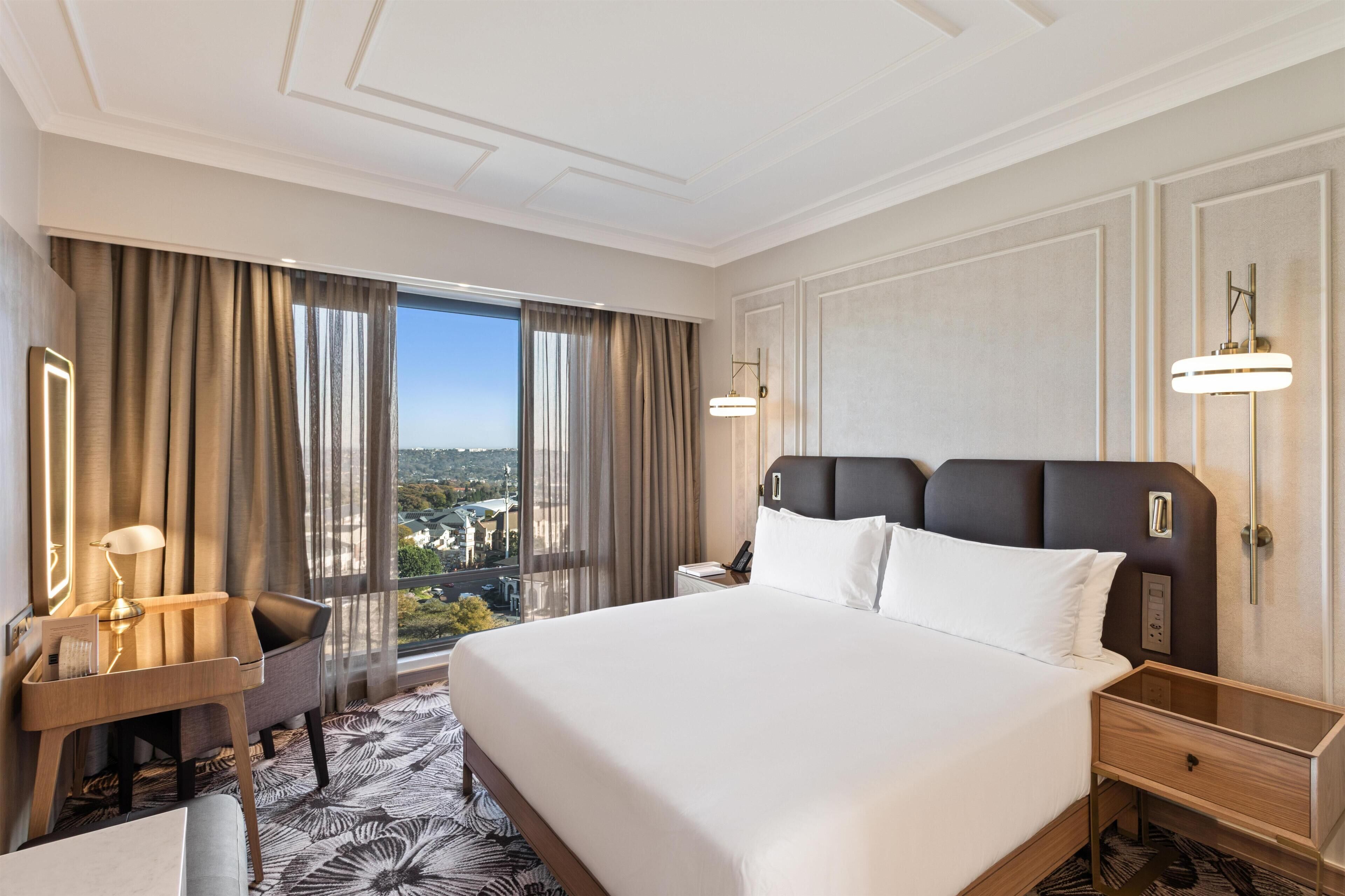 Quarto de hotel com uma cama grande, uma escrivaninha com abajur, telefone e vista para a cidade.