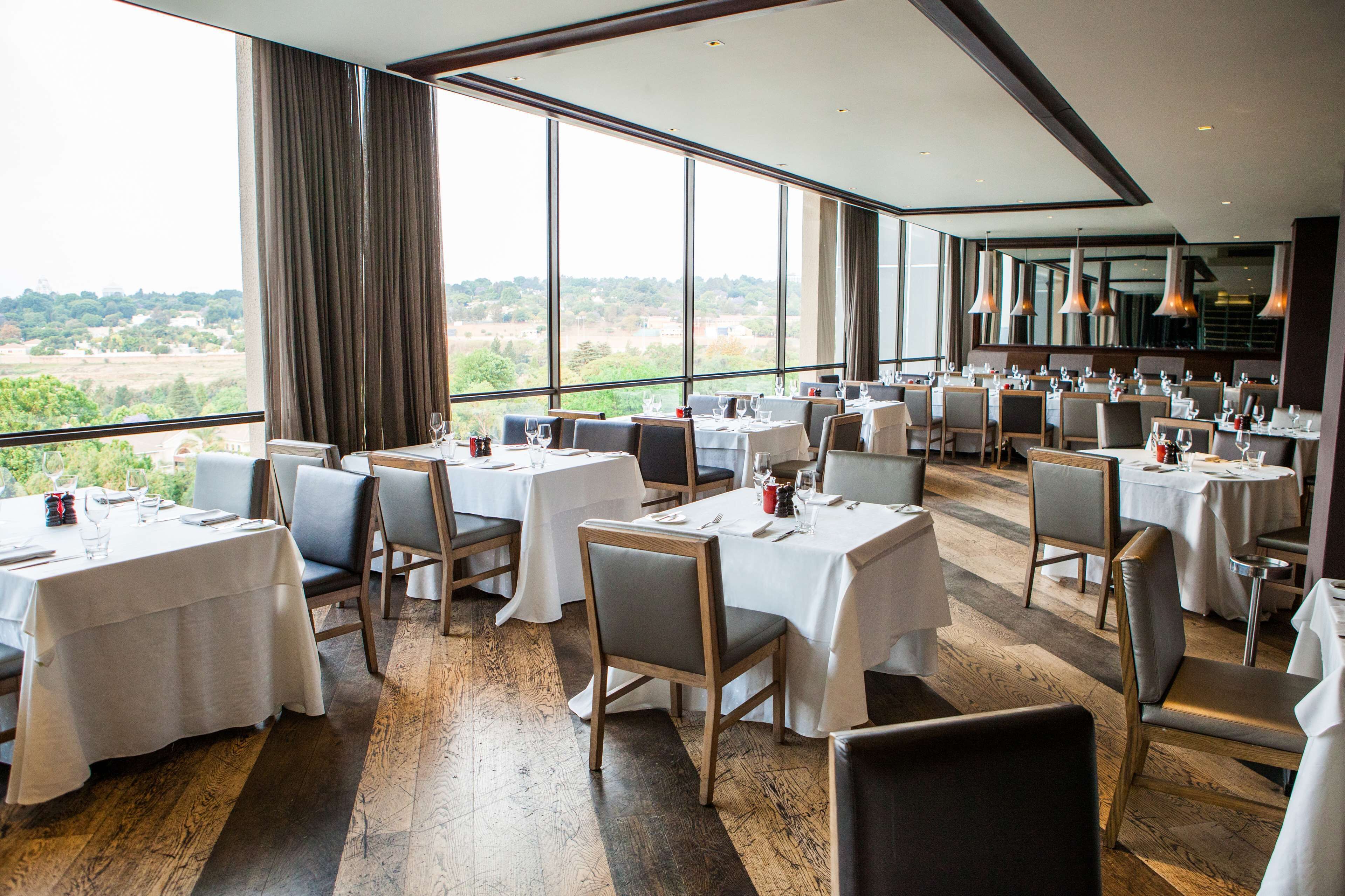 Um restaurante com várias mesas e cadeiras, grandes janelas proporcionando vista para o campo.