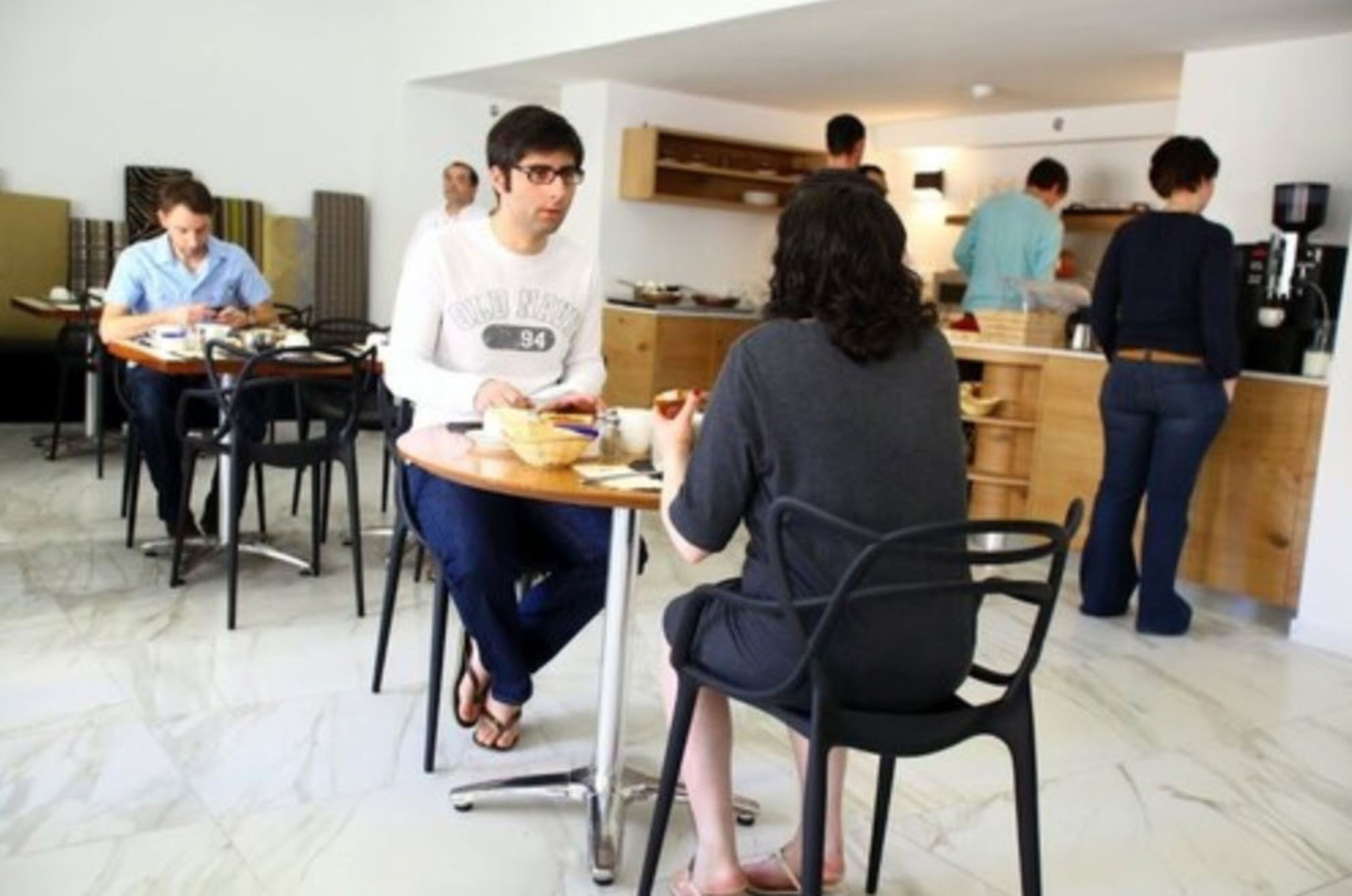Uma área de refeições compartilhada com mesas e cadeiras, uma cozinha com pessoas cozinhando e um design de interiores moderno.